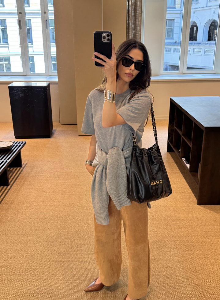 Woman wearing a casual gray t-shirt with a matching sweater tied at the waist%2C tan suede wide-leg pants%2C brown pointed-toe heels%2C black Chanel leather shoulder bag%2C and oversized black sunglasses in a modern indoor setting.